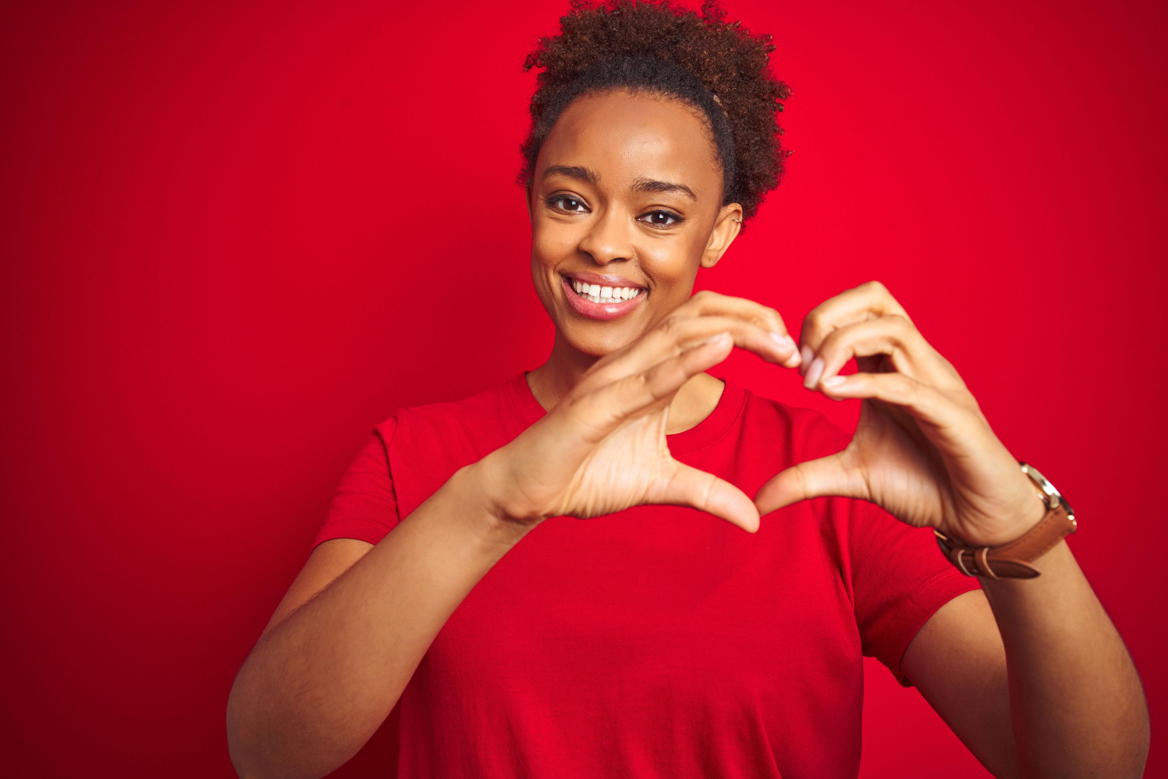 Women wearing red makes a heart with her hands against a red background.